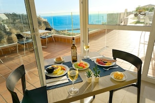Uma mesa de jantar com pratos de comida, uma garrafa de vinho e copos em uma varanda com vista para o mar.