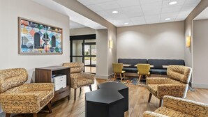 Um hall de hotel com poltronas estampadas, uma mesa de centro preta e um sofá. Há uma grande obra de arte emoldurada na parede e uma porta que dá para o exterior.