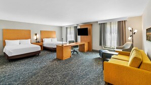 Quarto de hotel com duas camas, uma escrivaninha, um sofá, uma televisão e uma janela grande com cortinas.