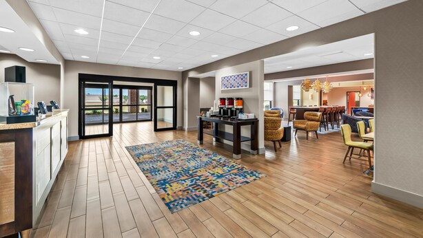 Um lobby moderno de hotel com balcão de recepção, espaço para café, área de refeições com cadeiras e mesas, e uma grande janela com vista.
