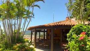 Uma área de estar coberta ao ar livre, com mesas e cadeiras de madeira, rodeada por vegetação exuberante e um céu azul límpido.
