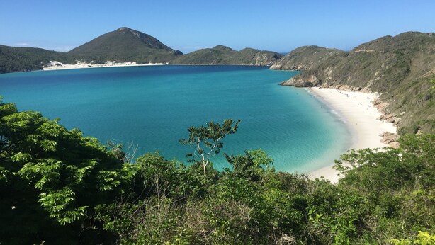 Paisagem costeira com mar azul cristalino, praia arenosa e colinas verdes exuberantes.