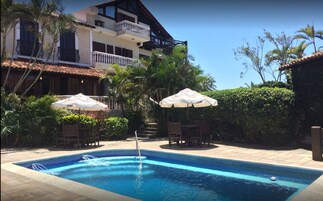 Piscina com dois guarda-sóis, um edifício com varanda e vegetação tropical.
