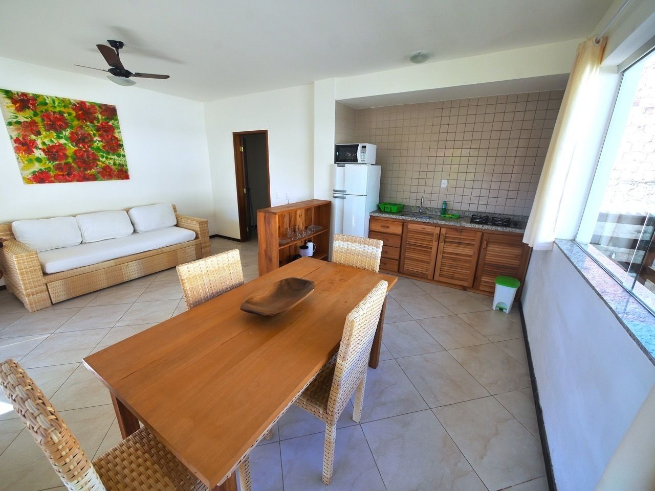 Uma sala de jantar com mesa de madeira e cadeiras de vime, uma cozinha com micro-ondas e armários, e um sofá na sala de estar.