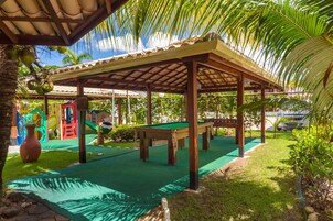Uma mesa de bilhar sob um pavilhão de madeira com feltro verde, rodeada por palmeiras e um espaço infantil.