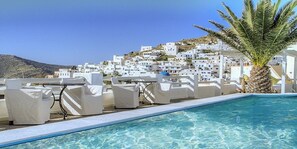 Área à beira da piscina com espreguiçadeiras brancas e uma palmeira, com vista para um morro de edifícios brancos.