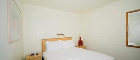 Quarto de hotel com uma cama de solteiro, mesa de cabeceira, espelho e um quadro pendurado na parede.