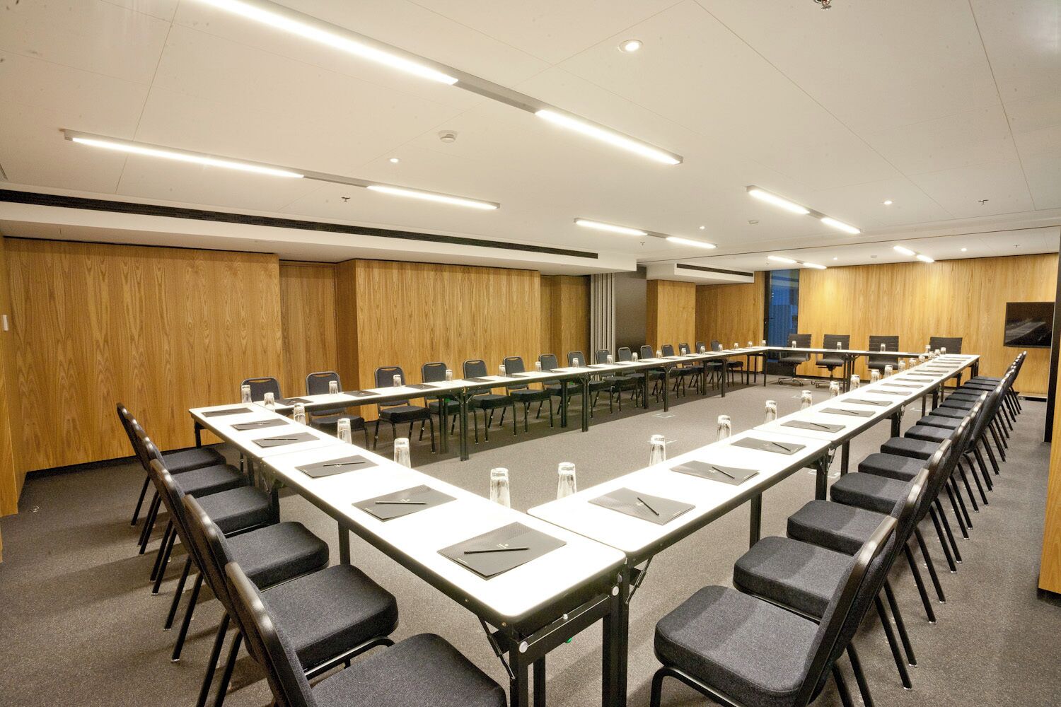 Uma sala de conferências com mesas e cadeiras compridas, painéis de madeira e iluminação embutida.