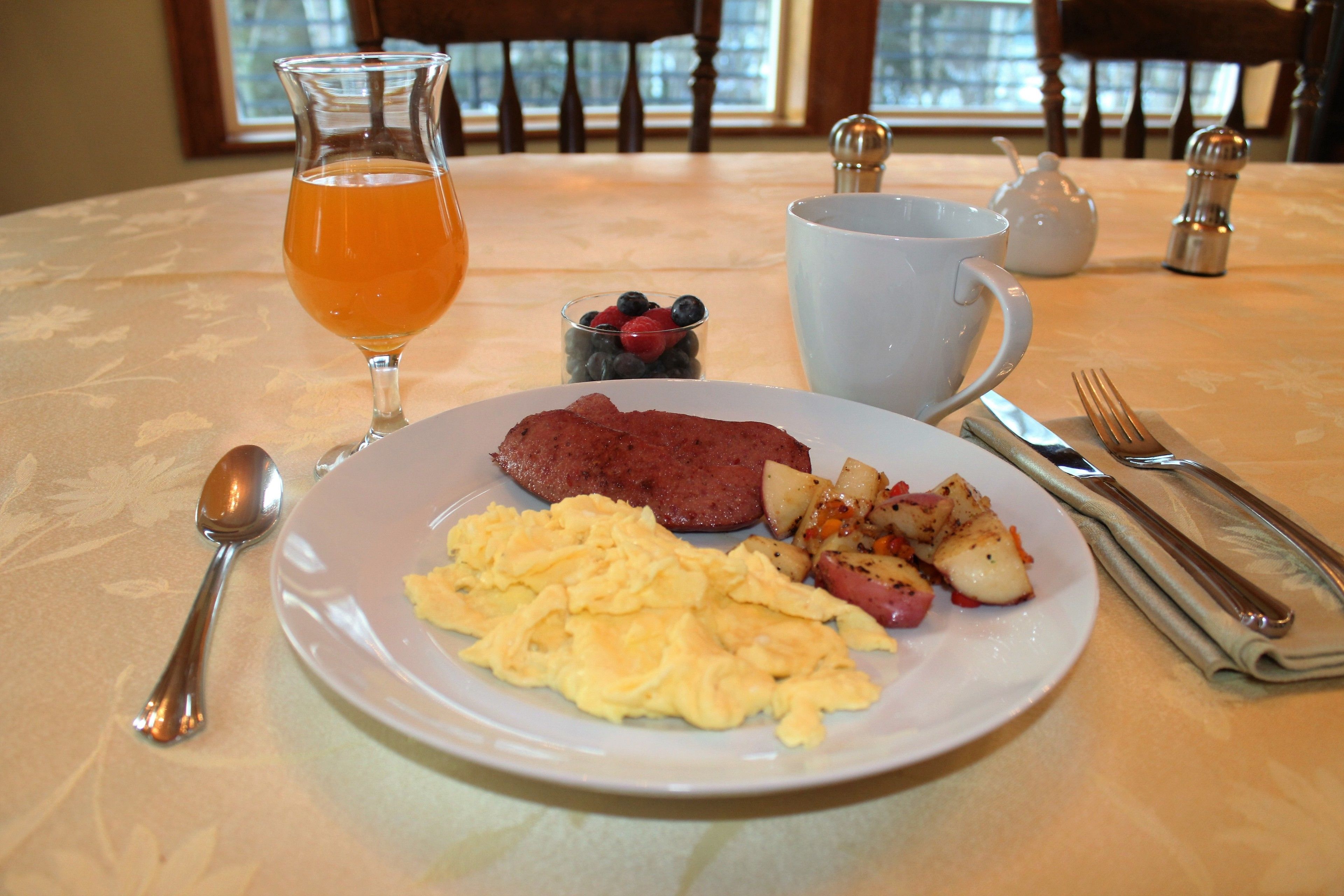 Prato de café da manhã com ovos mexidos, bacon e batatas, um copo de suco de laranja e uma pequena tigela de frutas vermelhas sobre uma mesa com uma caneca branca e saleiro e pimenteiro.
