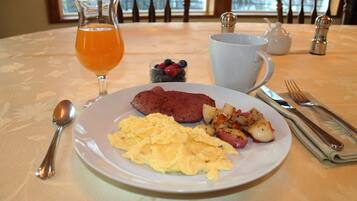 Prato de café da manhã com ovos mexidos, bacon e batatas, um copo de suco de laranja e uma pequena tigela de frutas vermelhas sobre uma mesa com uma caneca branca e saleiro e pimenteiro.
