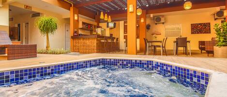 Uma área de hidromassagem com piscina azulejada, assentos de madeira e um bar com balcão e cadeiras.