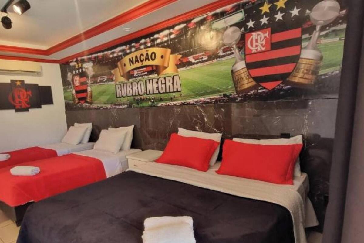 Quarto de hotel com duas camas, roupa de cama vermelha, um mural grande de um estádio de futebol e um logotipo na parede.