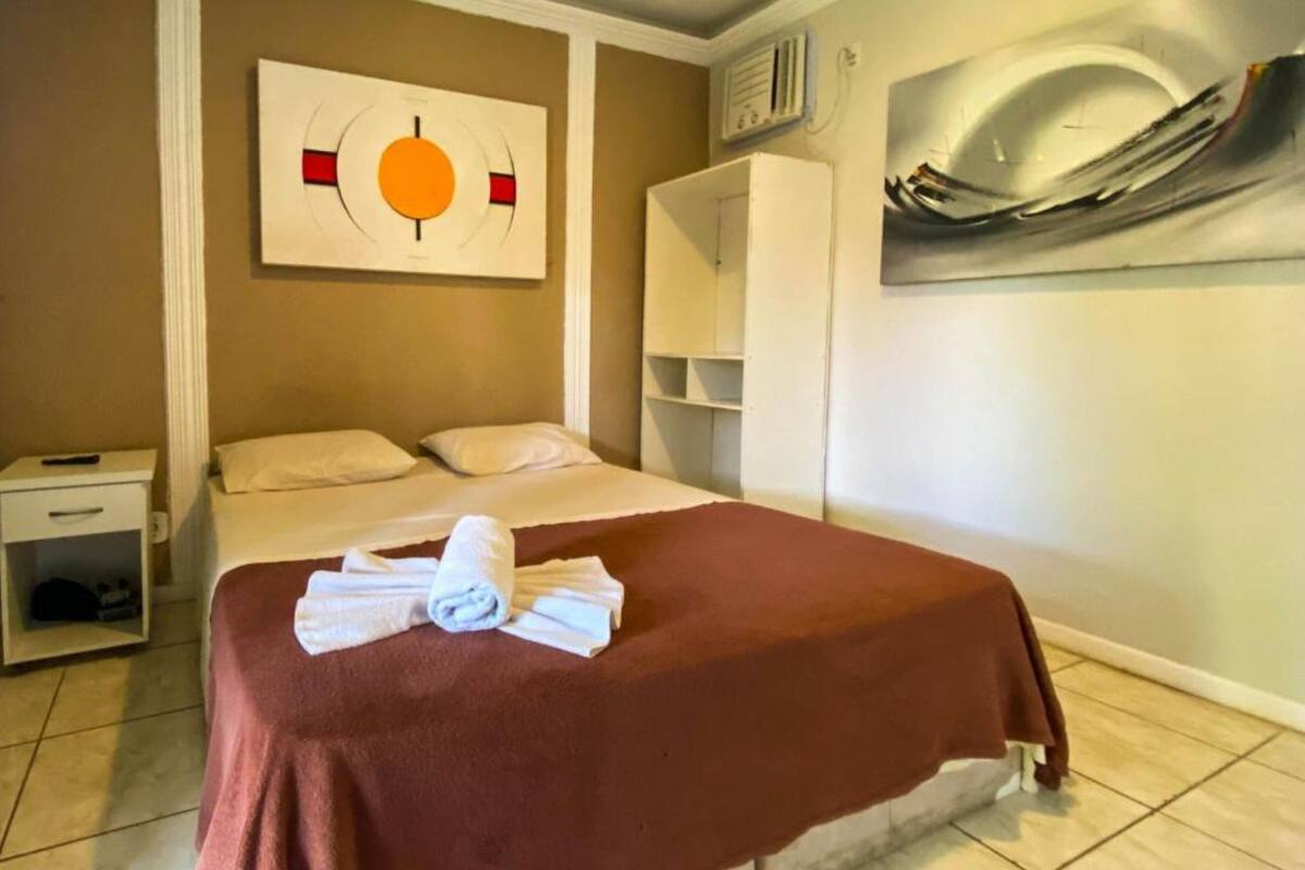 Quarto de hotel com uma cama, uma mesinha de cabeceira branca, um guarda-roupa branco e um quadro na parede.