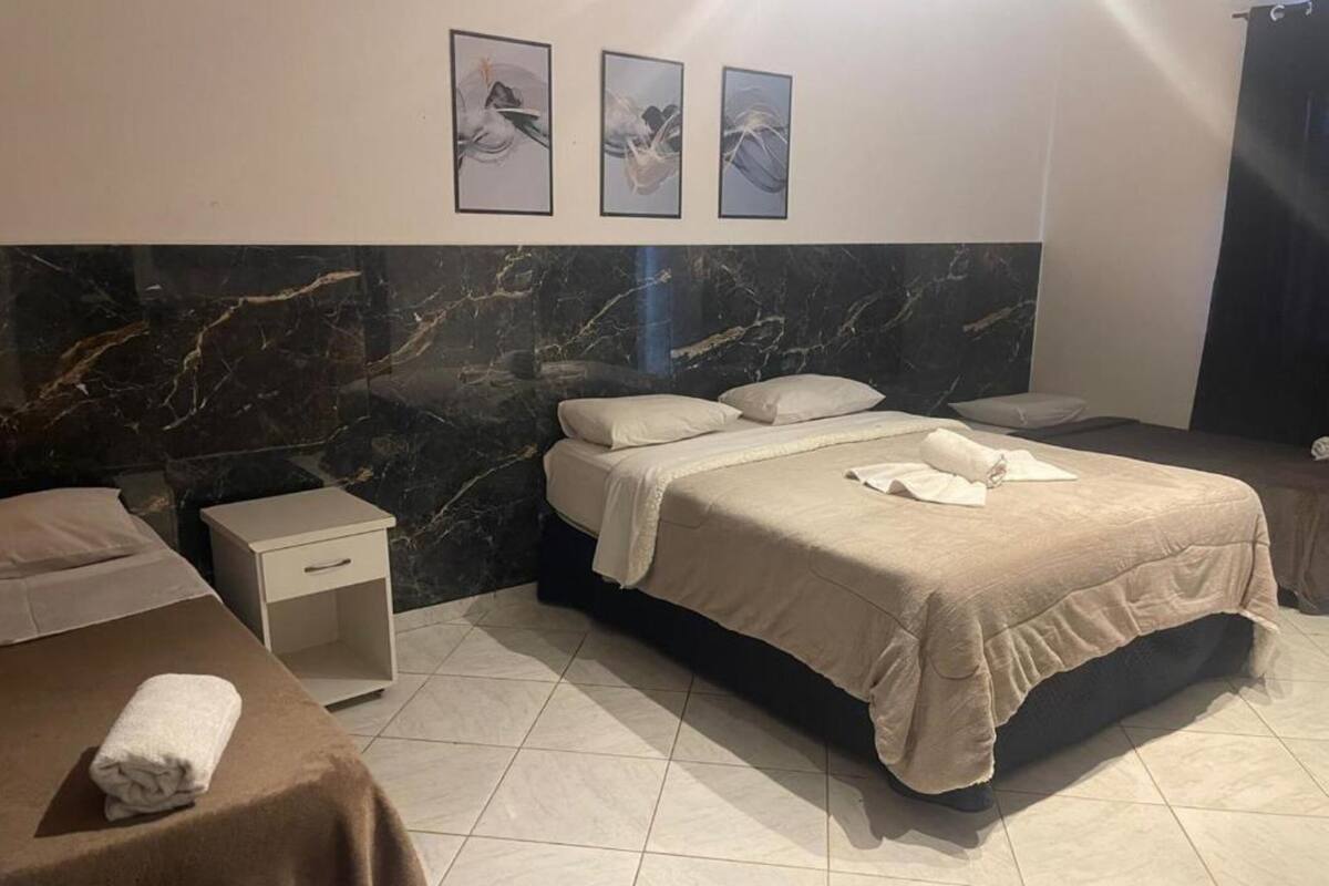 Quarto de hotel com uma cama grande, duas mesas de cabeceira e uma cabeceira de mármore. Há três quadros na parede acima da cama.