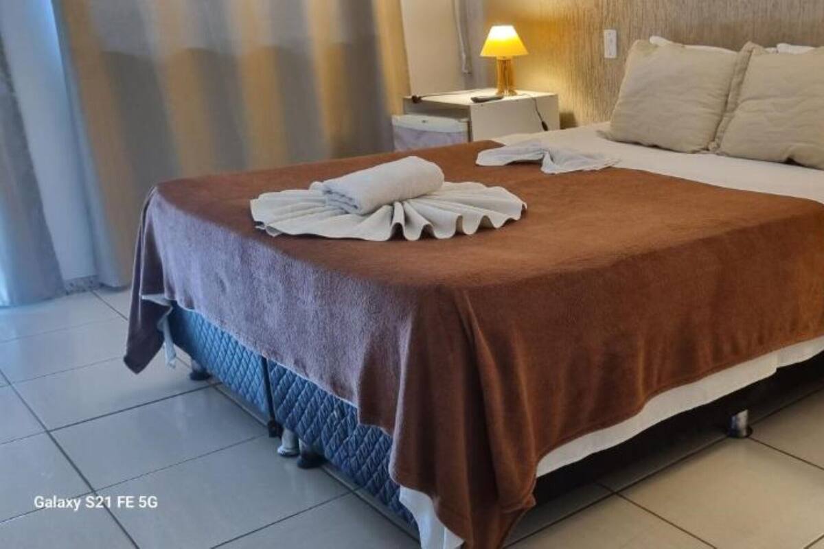 Quarto de hotel com uma cama, duas mesas de cabeceira com abajures, um criado-mudo e piso ladrilhado.