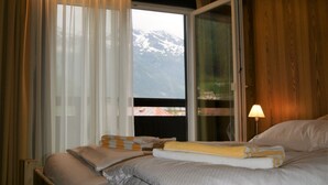 Uma cama com estrutura de madeira e roupa de cama branca, uma janela com cortinas e vista para montanhas cobertas de neve.