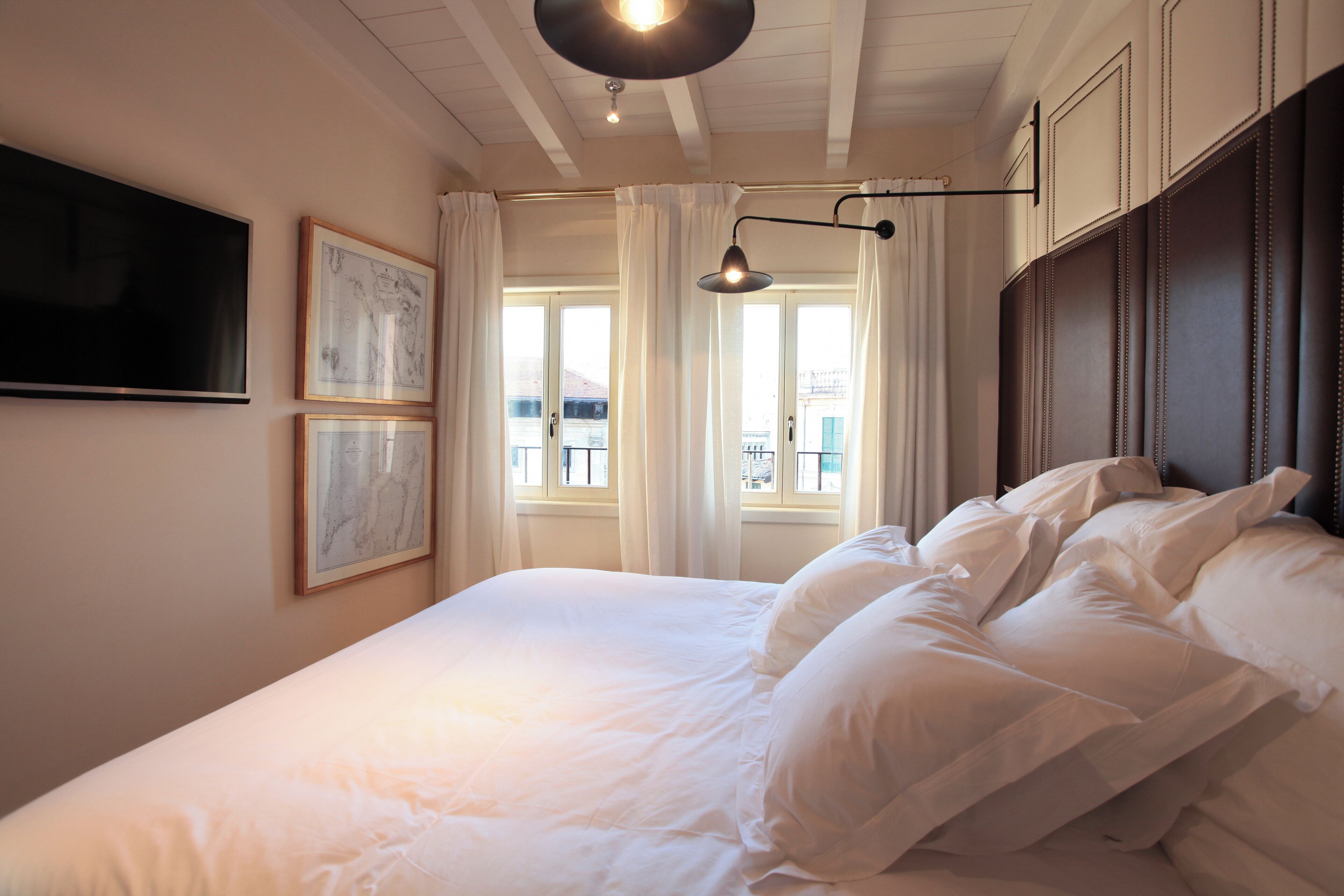Cama bem arrumada com roupa de cama branca em quarto com cabeceira de madeira, luminária no teto e duas janelas com cortinas.