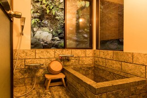 Uma casa de banho tradicional japonesa com uma banheira de pedra, um banquinho de madeira e uma janela com vista para um jardim rochoso.