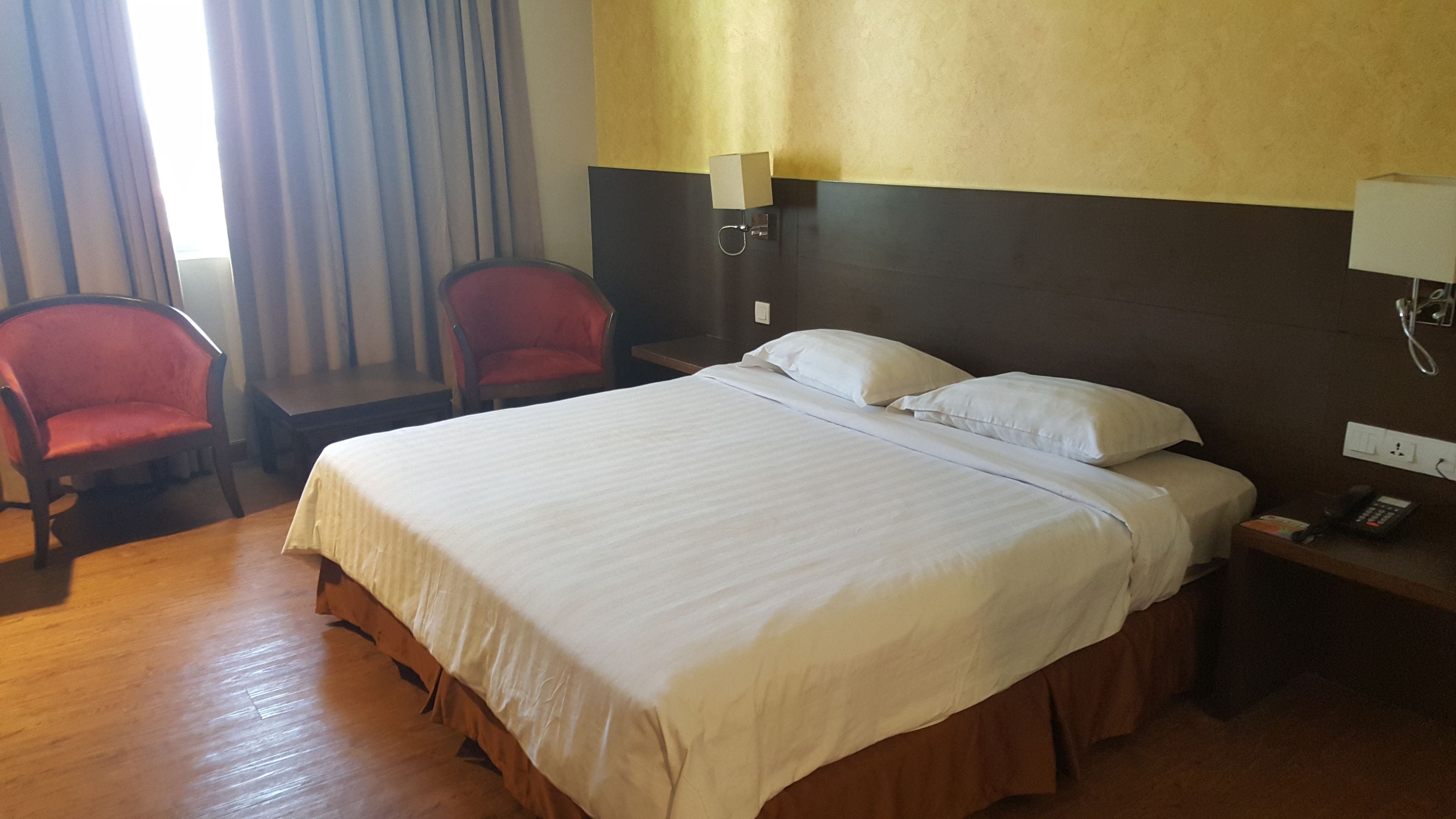 Quarto de hotel com uma cama grande, duas cadeiras vermelhas, uma mesa de madeira, uma mesinha de cabeceira com abajur e um telefone.