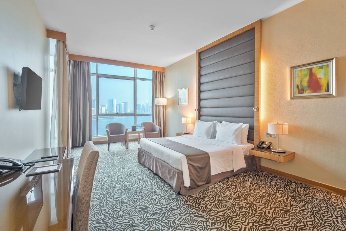 Quarto de hotel com uma cama grande, escrivaninha com telefone, cadeira, abajur, mesa de cabeceira com luminária e vista para a cidade através de amplas janelas.