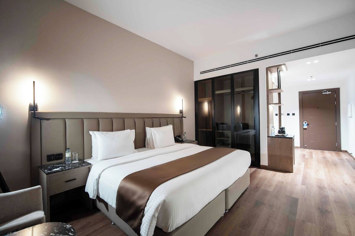 Quarto de hotel moderno com uma cama grande, mesa de cabeceira, uma cadeira e um guarda-roupa.