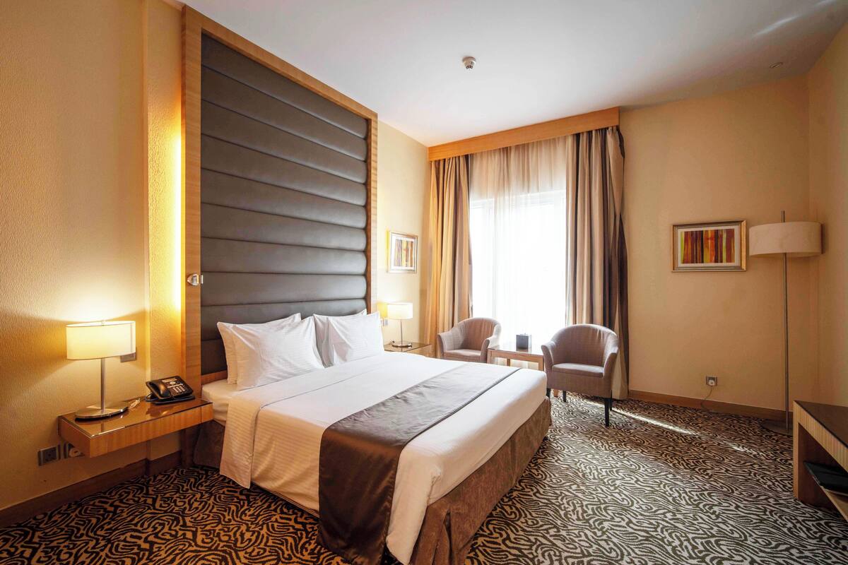 Quarto de hotel com uma cama grande, uma escrivaninha, uma cadeira, um abajur e uma janela com cortinas.