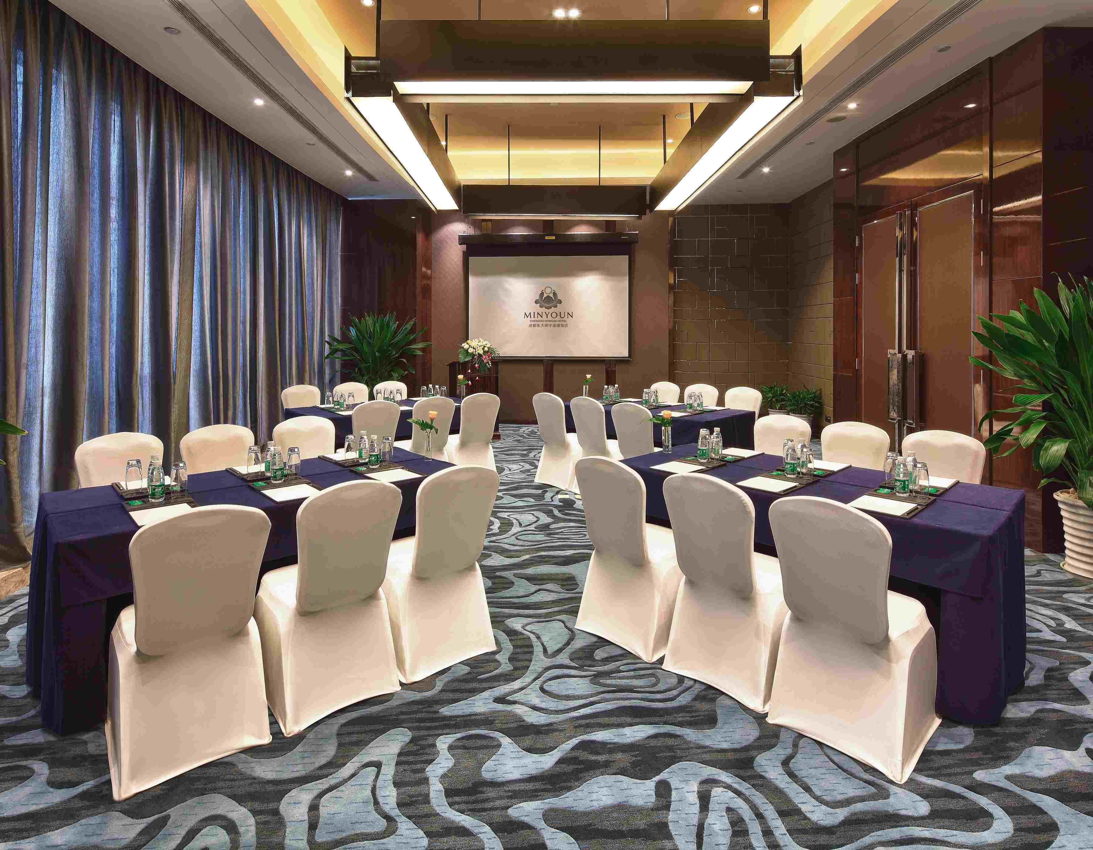 Uma sala de conferência com fileiras de mesas e cadeiras, um púlpito e uma tela exibindo o logotipo.