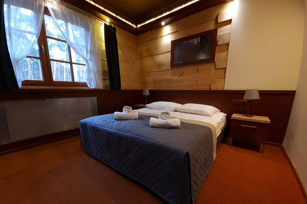 Quarto de hotel com uma cama grande, uma janela com cortinas, um quadro na parede e uma mesa de cabeceira com abajur.