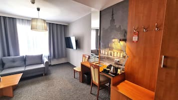 Quarto de hotel com sofá, mesa de jantar com cadeiras, televisão na parede e pintura de uma paisagem urbana.