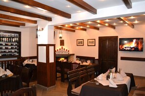 Interior de restaurante com vigas de madeira, lareira, mesas de jantar preparadas para hóspedes e uma televisão fixada na parede.