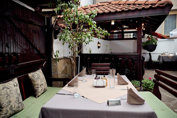 Uma área de refeições com mesa posta para dois, um banco e uma pérgula com uma árvore.