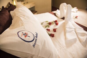 Uma cama bem arrumada com travesseiro branco exibindo um logotipo e texto, e rosas vermelhas espalhadas sobre a cama.