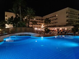 Uma piscina com design curvo, rodeada por uma vedação de madeira. Ao fundo, um edifício de vários andares com varandas e área de bar.