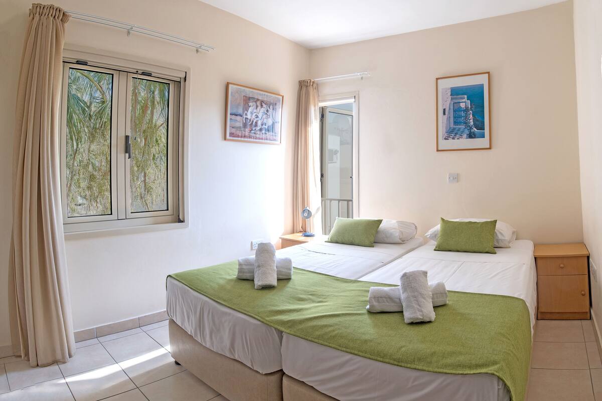 Quarto de hotel com duas camas, uma janela com cortinas, uma mesa de cabeceira e duas molduras na parede.