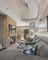 Uma sala de estar moderna com parede de pedra, lareira, televisão de tela plana, sofá com almofadas florais e um espaço de jantar com cadeiras e mesa.