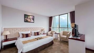 Quarto de hotel com uma cama grande, duas mesas de cabeceira, uma escrivaninha com um computador, uma cadeira e vista para um horizonte urbano através de grandes janelas.