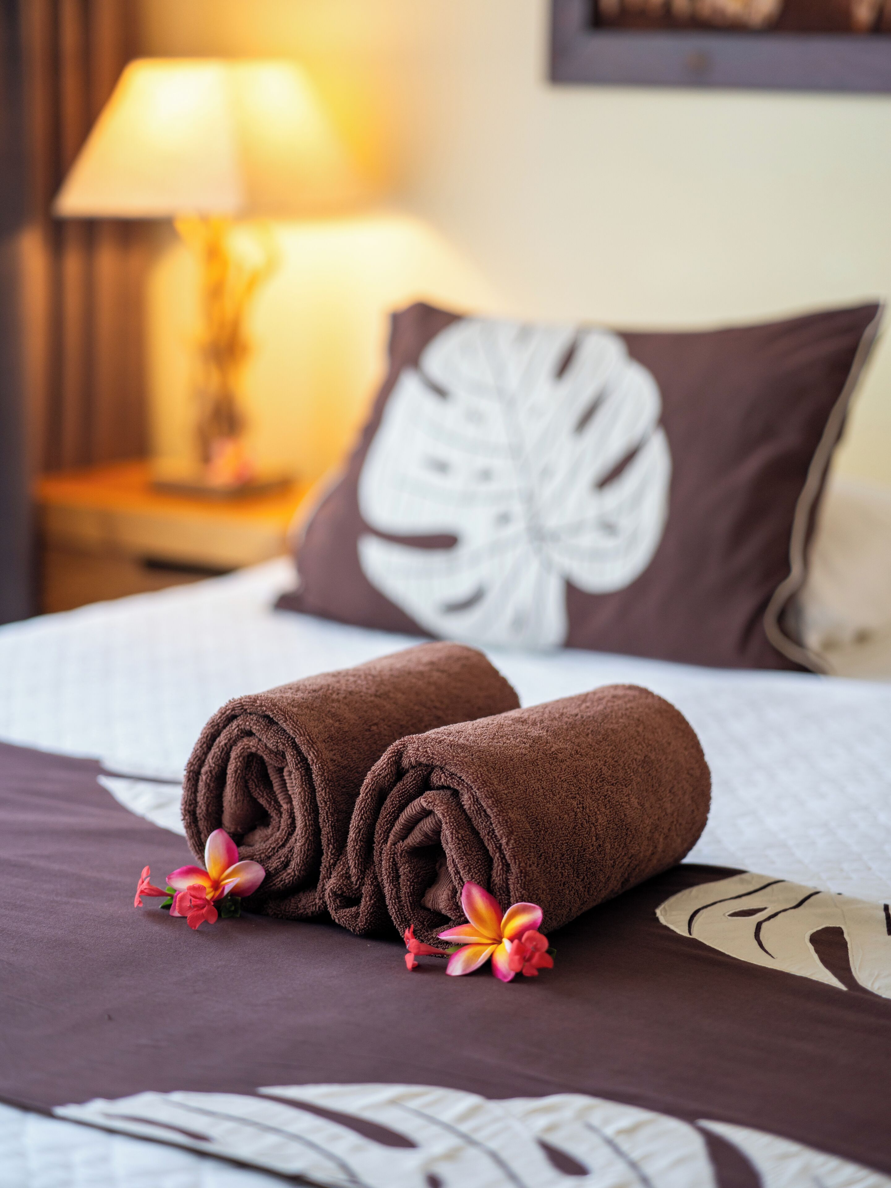 Cama bem arrumada com toalhas castanhas e travesseiro com folha tropical.
