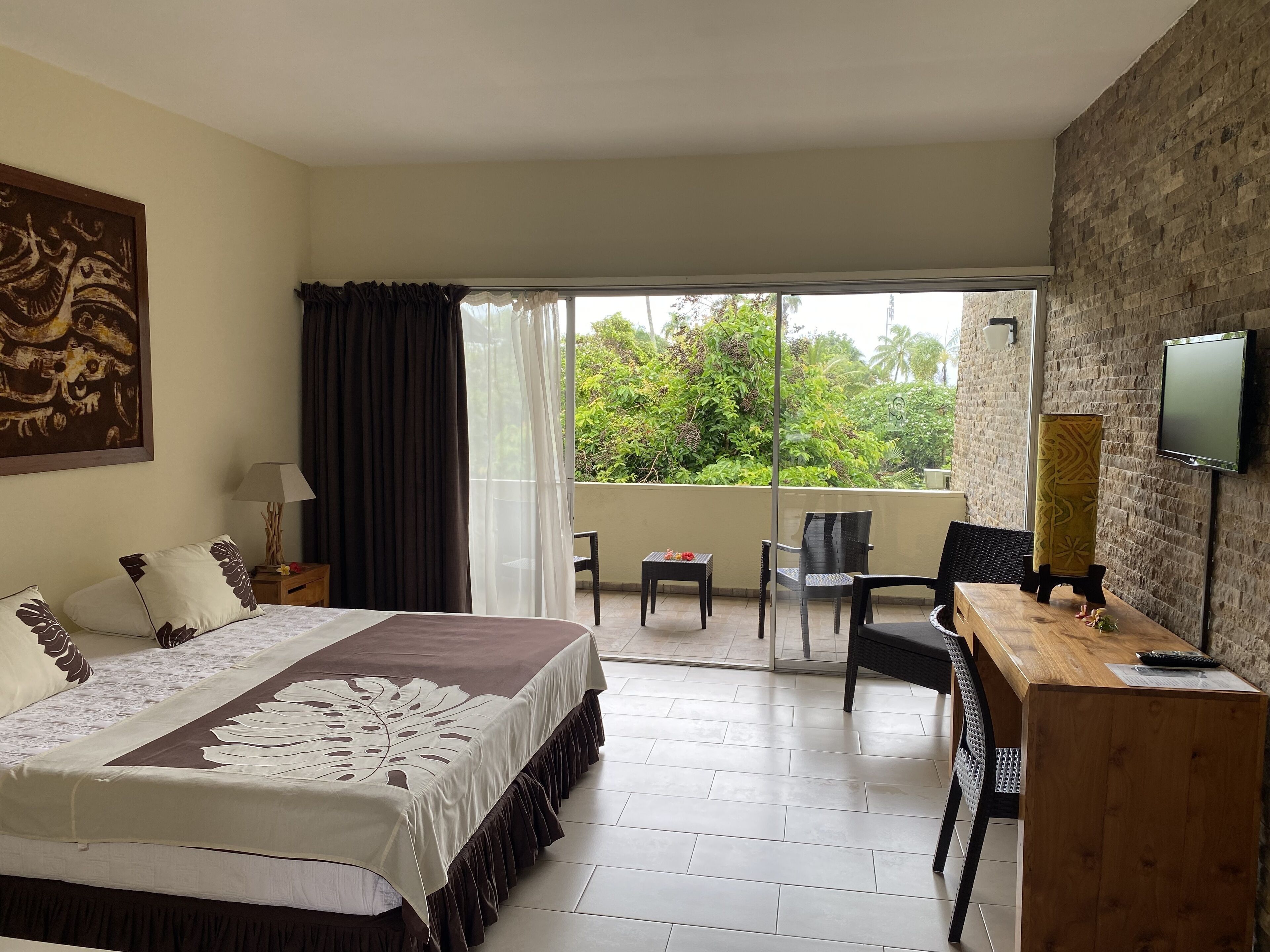 Quarto de hotel com uma cama, uma escrivaninha, uma cadeira, televisão e varanda com vista para o verde.