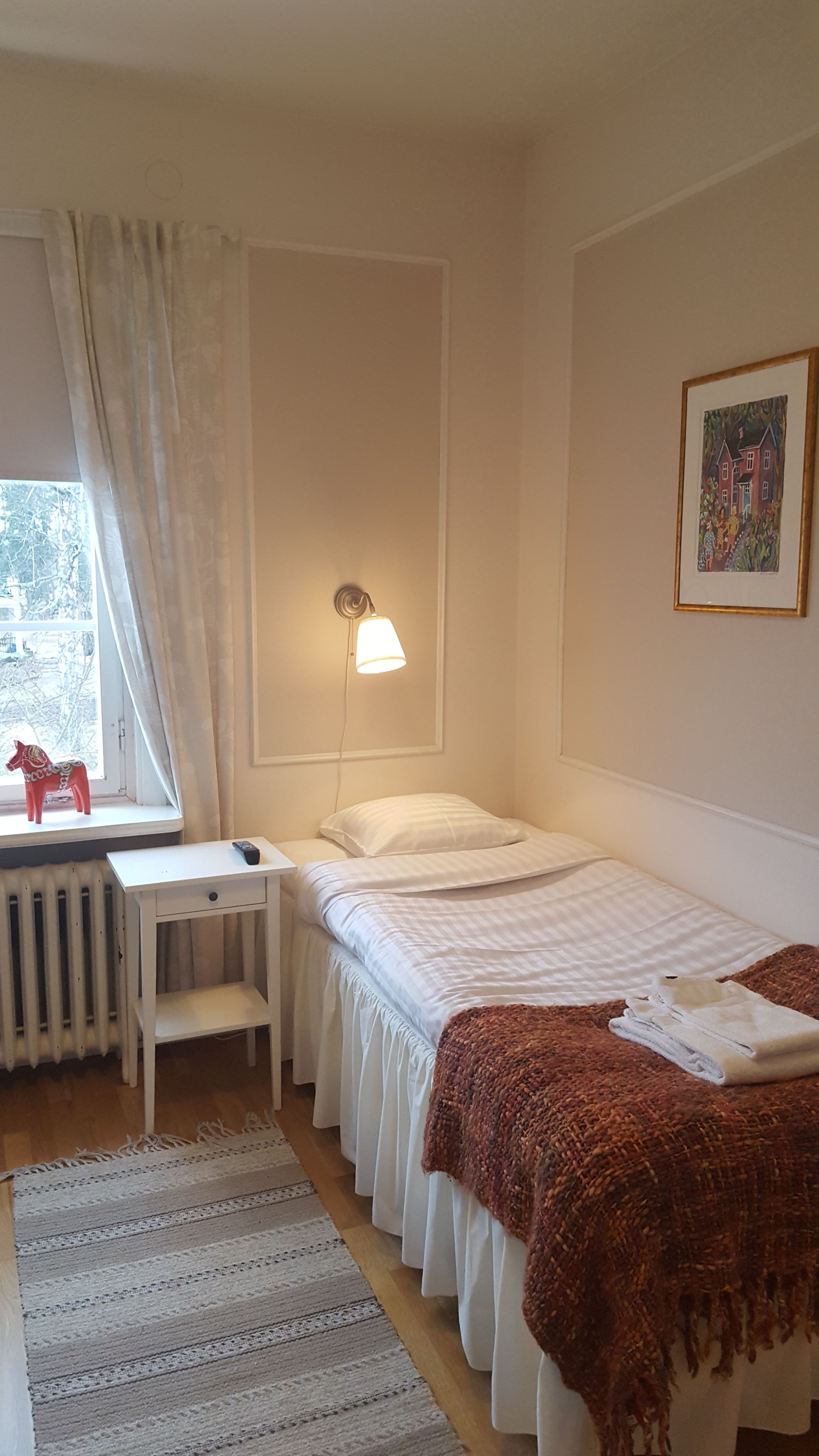 Quarto com uma cama de solteiro, uma escrivaninha, um radiador, uma janela com cortinas, uma luminária na parede e um quadro emoldurado.