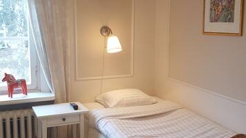 Quarto com uma cama de solteiro, uma escrivaninha, um radiador, uma janela com cortinas, uma luminária na parede e um quadro emoldurado.