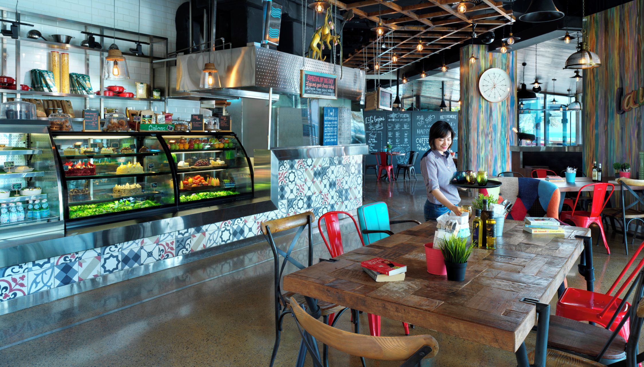 Interior de restaurante com balcão exibindo comida, mesa de madeira com livros e planta, e cadeiras vermelhas.