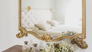 Quarto com um espelho grande e ornamentado, cômoda de madeira com objetos decorativos e cama com roupa de cama branca.