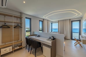 Quarto de hotel moderno com escrivaninha, cadeiras, minibar e vista para o mar.