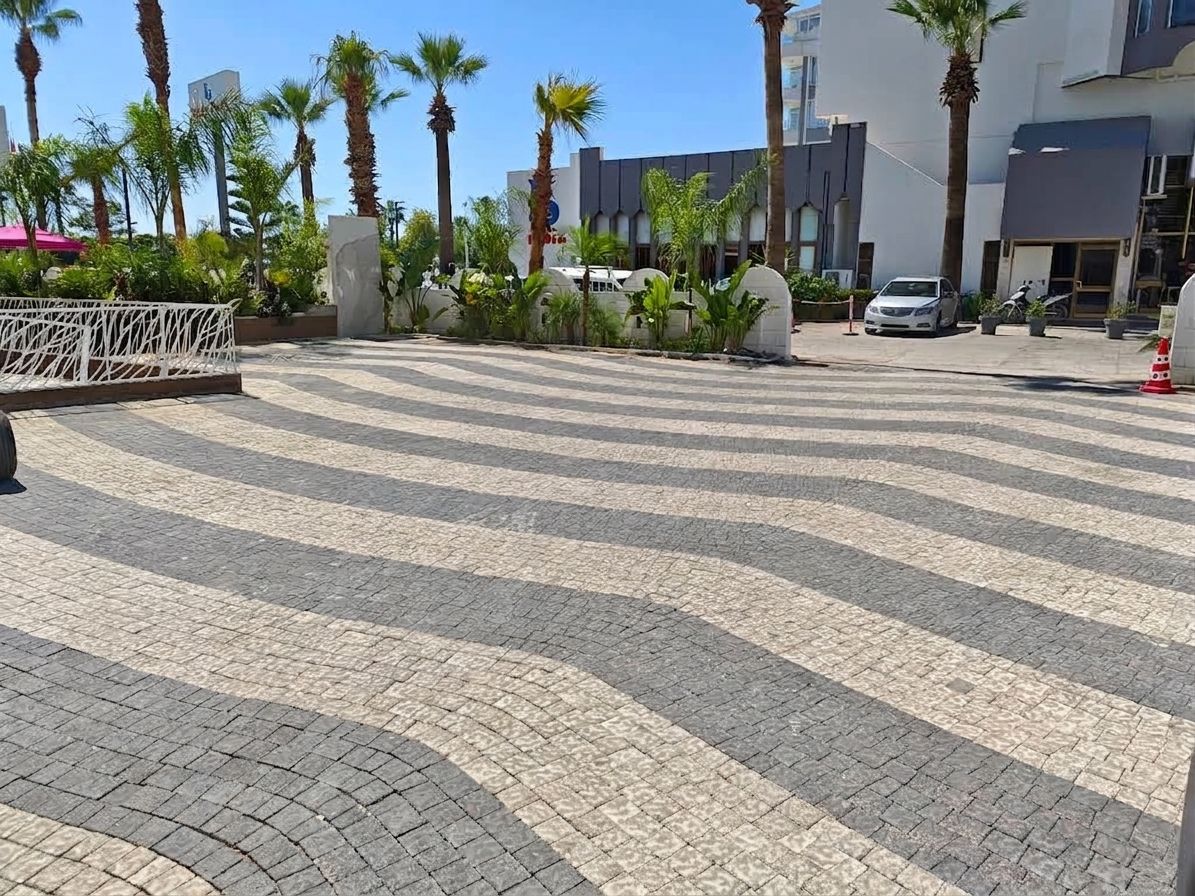 Uma área externa pavimentada com cerca branca, palmeiras e um edifício com design moderno ao fundo.