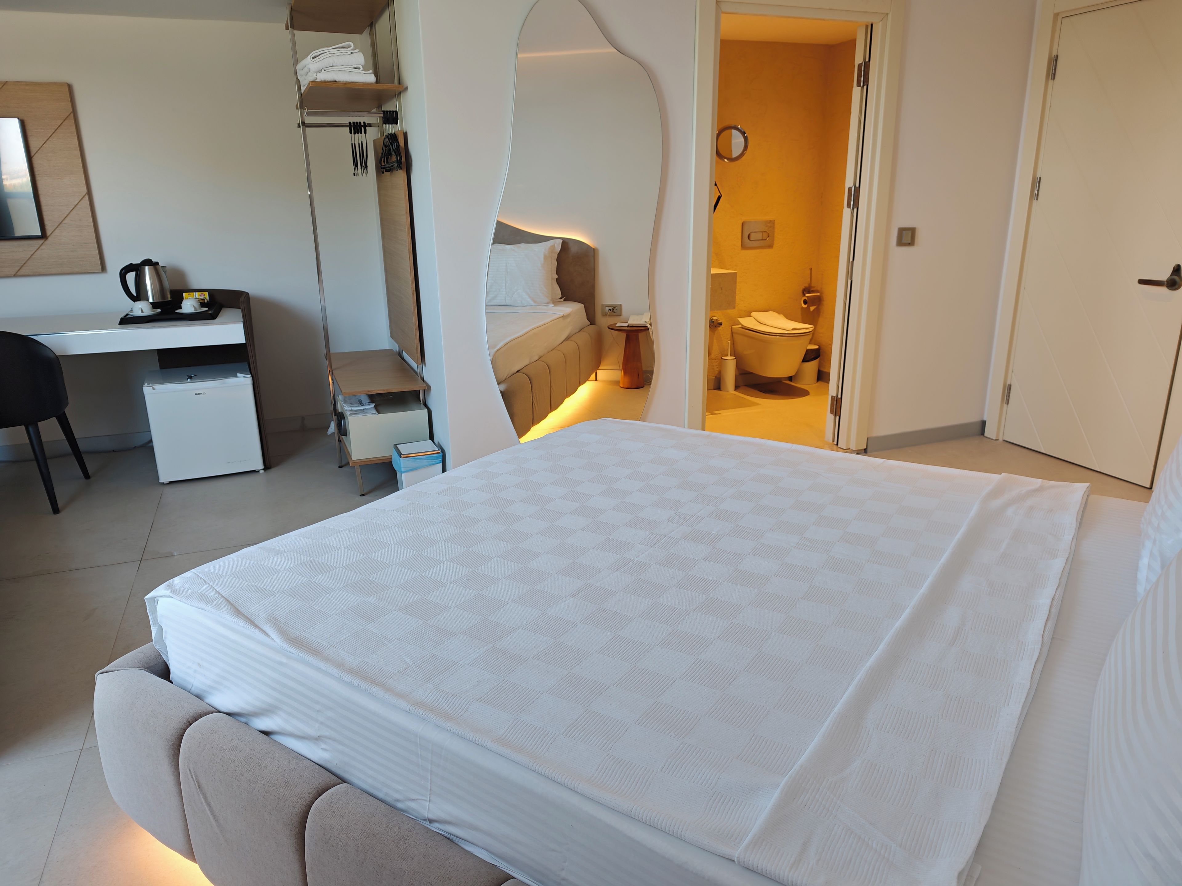 Quarto de hotel moderno com cama, escrivaninha, cadeira, pequeno frigorífico e casa de banho visível através de uma porta aberta.