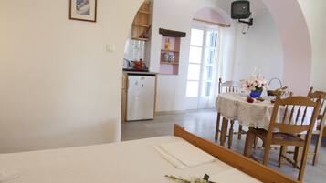 Quarto com cama, uma cadeira de madeira, mesa de jantar com cadeiras, cozinha ao fundo e um corredor.