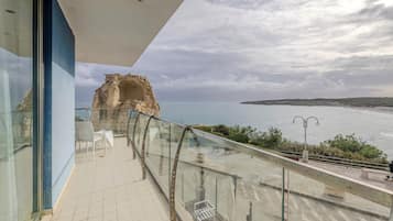 Varanda com guarda-corpo de vidro com vista para área costeira com ruínas e o mar.