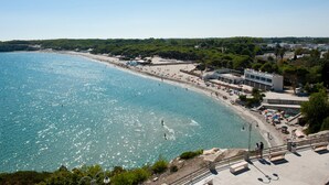 Uma praia costeira com águas azuis cristalinas, areal arenoso e alguns edifícios ao fundo.