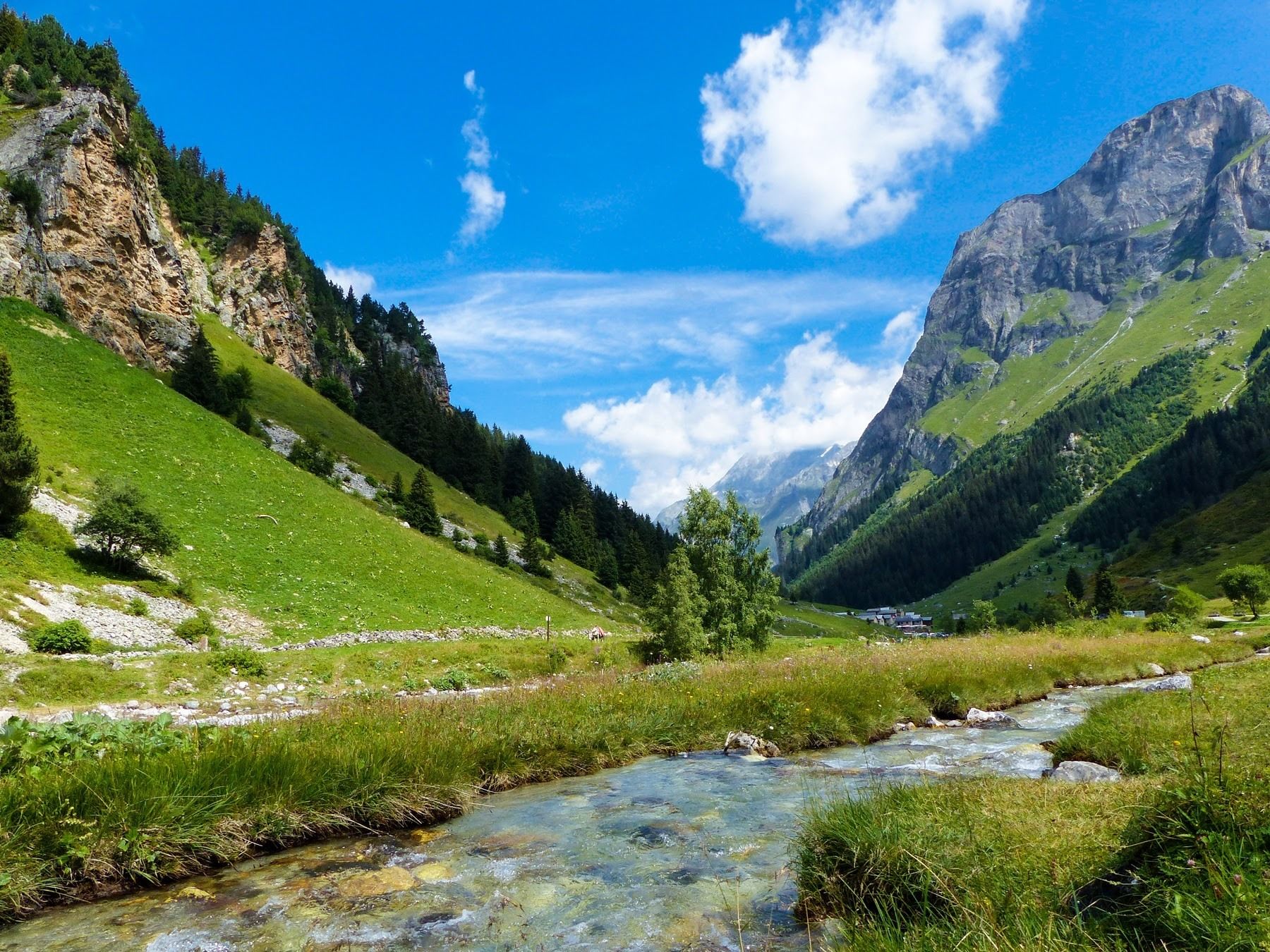 Paisagem montanhosa com um riacho límpido, campos verdejantes e penhascos rochosos.
