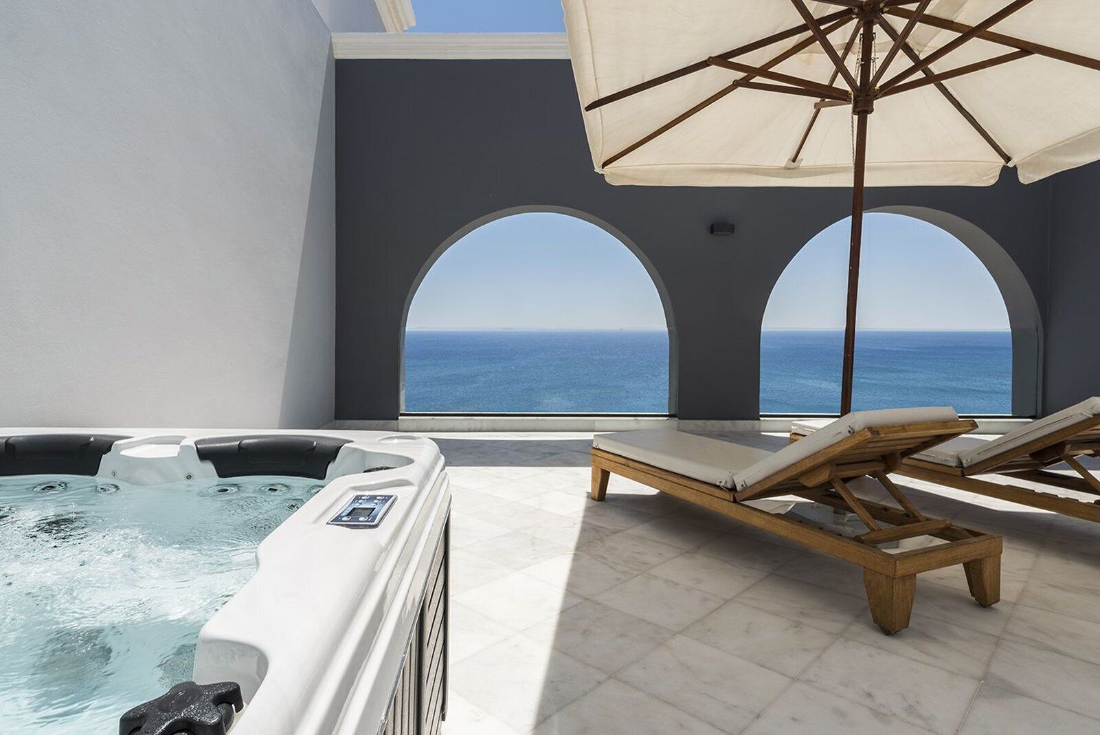 Uma banheira de hidromassagem com painel de controle, duas cadeiras de descanso e um guarda-sol numa varanda com vista para o oceano.